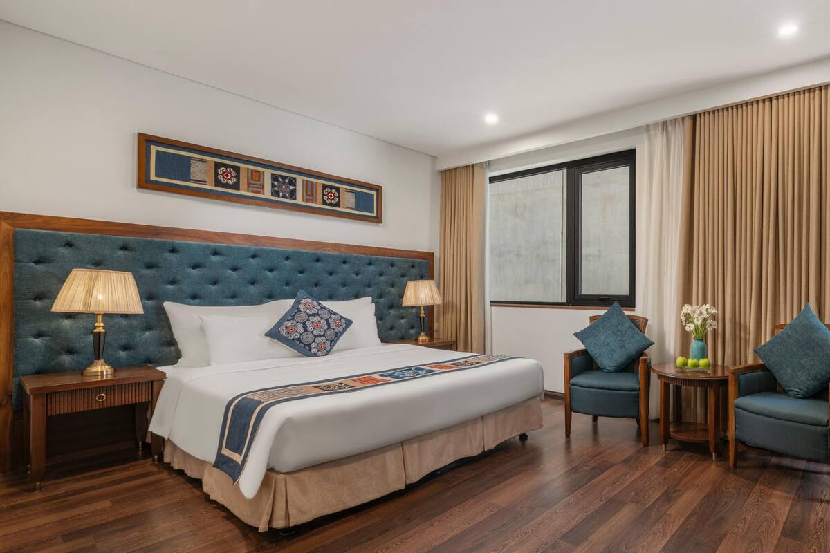 Quarto de hotel com uma cama grande, duas mesas de cabeceira com luminárias, uma cadeira, uma mesinha com flores e uma janela com cortinas.