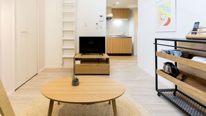 Uma sala de estar moderna com televisão, móveis de madeira, uma pequena planta e uma área de cozinha ao fundo.