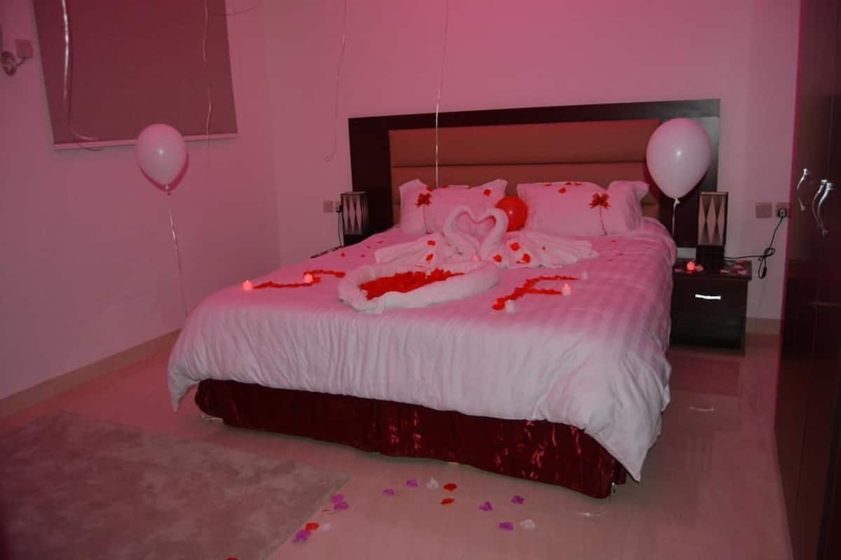 Cama bem arrumada com edredom branco e detalhes vermelhos, duas luminárias de cabeceira e um relógio fixado na parede.