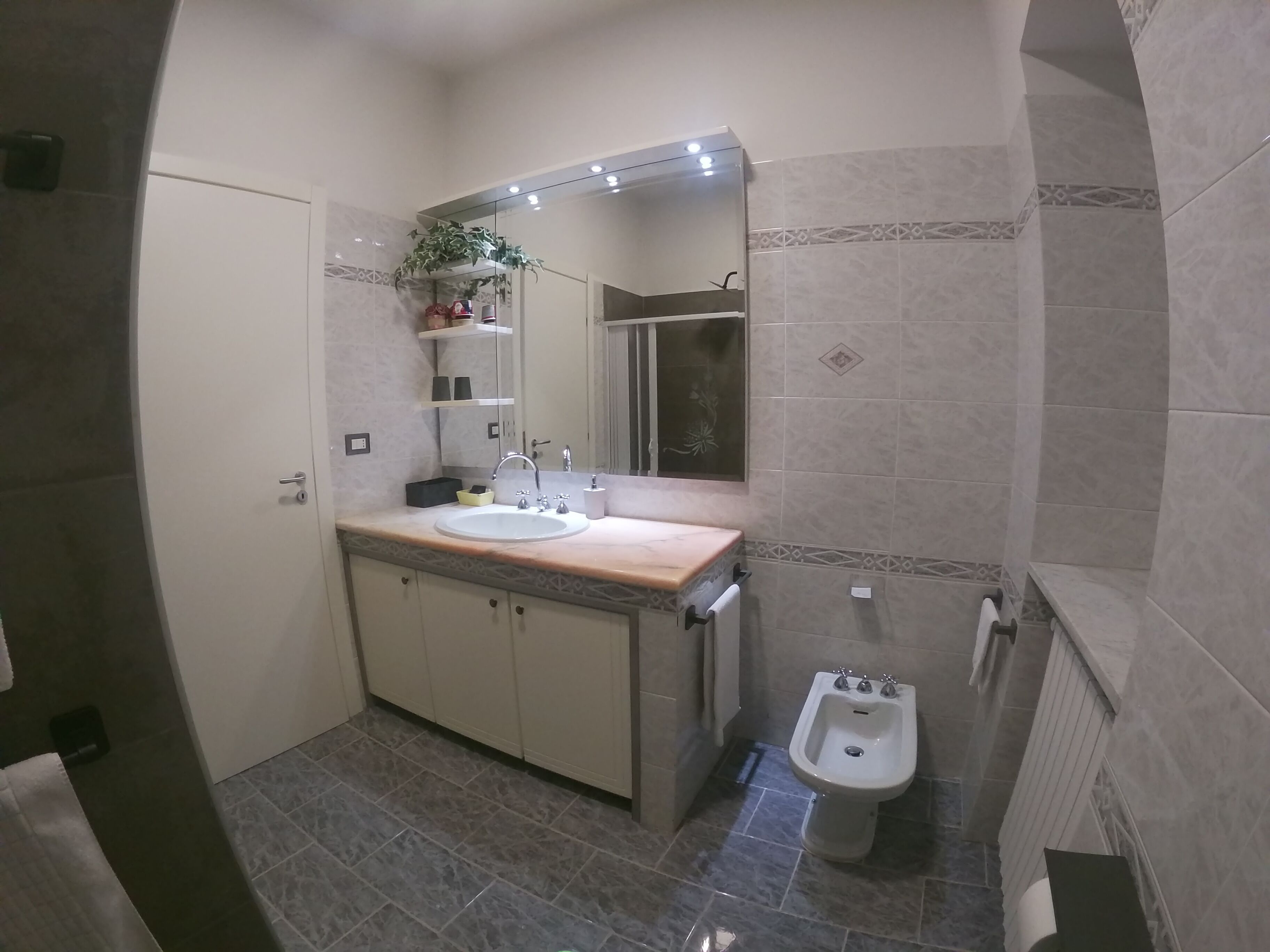 Um banheiro moderno com pia, espelho e bidê. Possui piso em azulejos e uma porta que dá para outro cômodo.