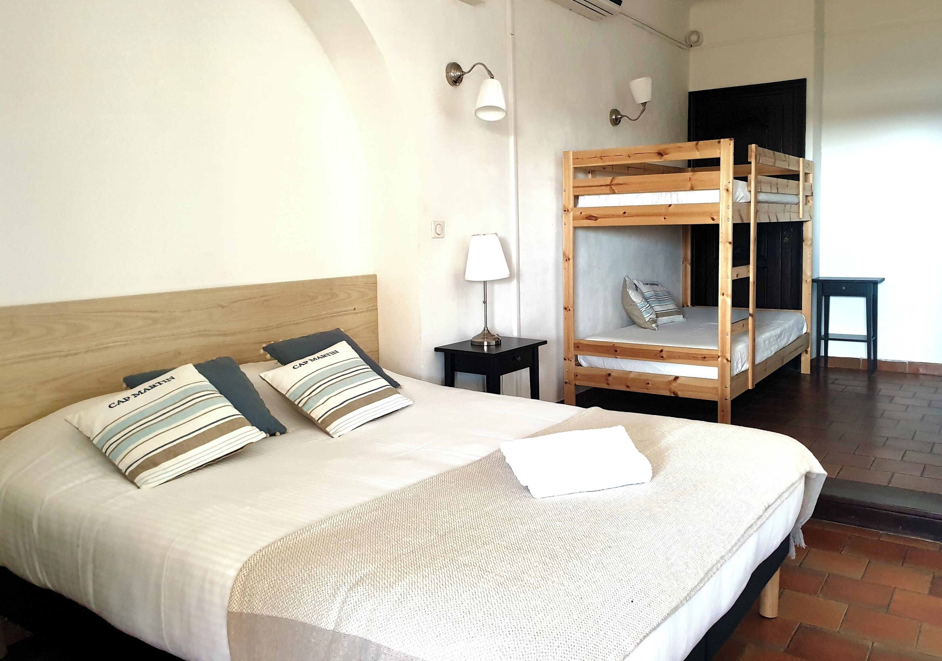 Quarto de hotel com uma cama grande, duas almofadas, uma luminária de cabeceira, uma beliche de madeira e uma mesa pequena.