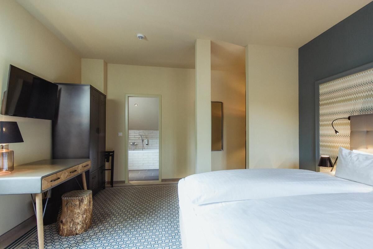 Quarto de hotel com cama, escrivaninha com luminária, televisão e banheiro visível por uma porta aberta.