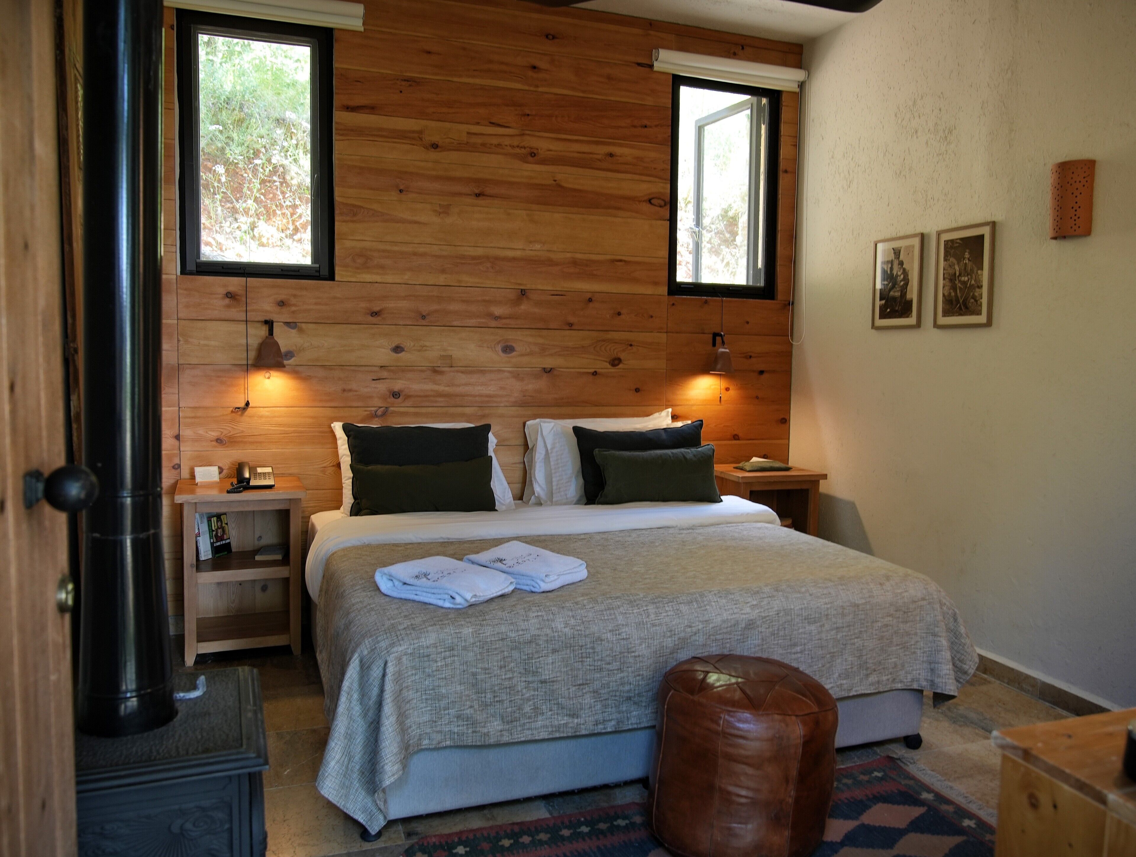 Quarto com cabeceira de madeira, duas luminárias de apoio, cama com travesseiros, mesa de cabeceira, uma pequena mesa, um banquinho e uma janela com vista para o verde.
