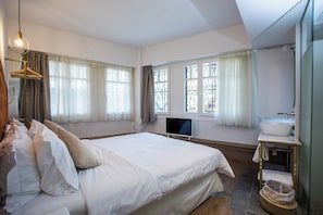 Quarto de hotel com cama, televisão, banheiro com pia e amplas janelas com cortinas.