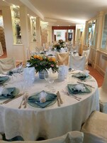 Uma sala de jantar formal com mesas redondas preparadas para uma refeição, com toalhas de mesa verdes, cadeiras brancas e arranjos florais.