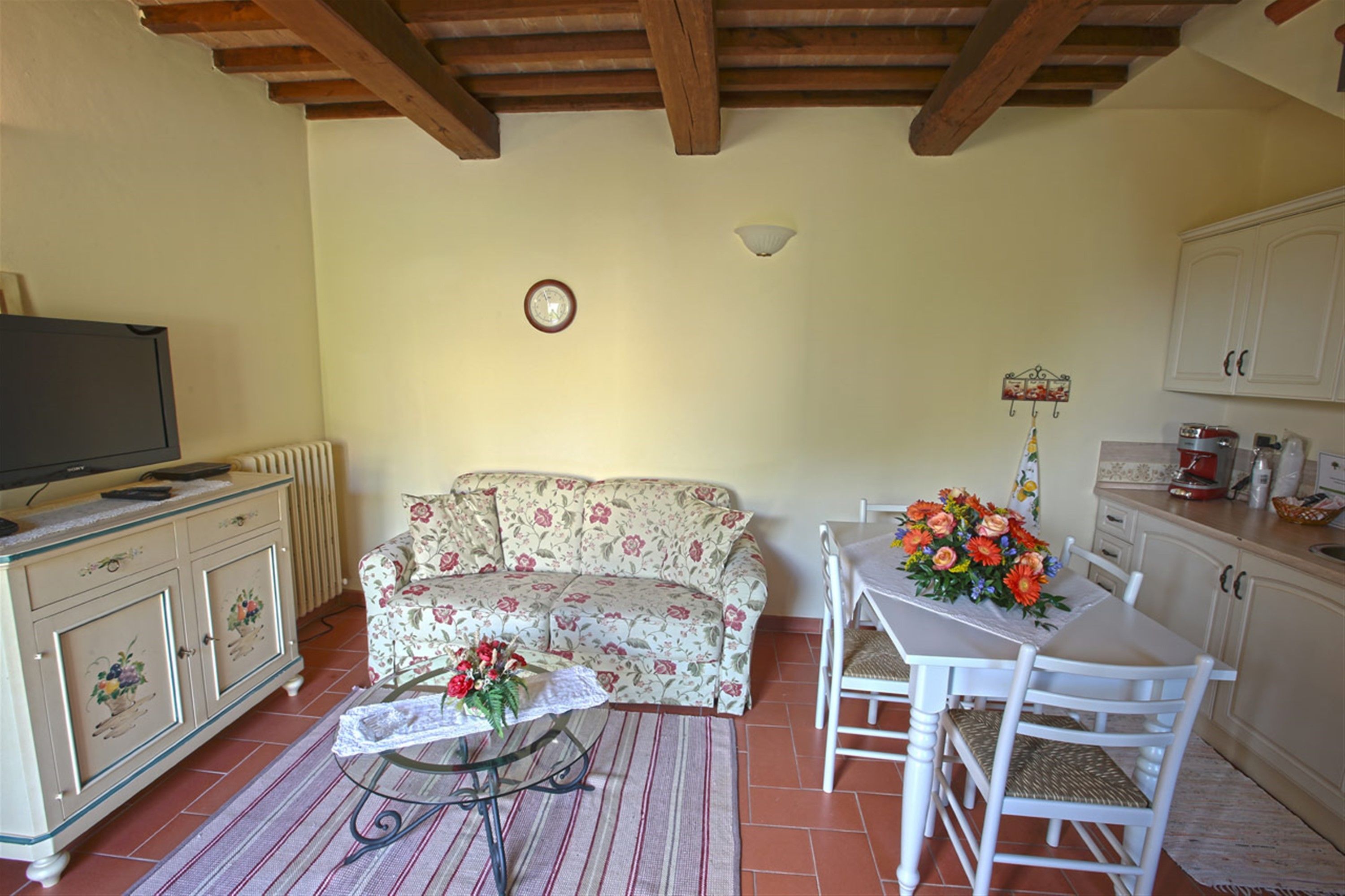Uma sala de estar acolhedora com um sofá floral, uma mesa de jantar com cadeiras, uma televisão sobre um móvel e uma cozinha com armários e eletrodomésticos.