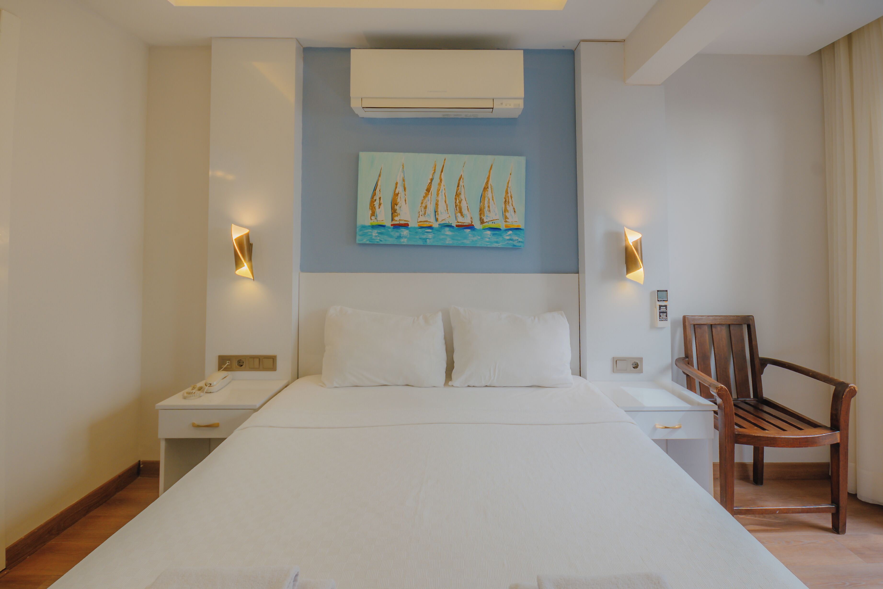 Quarto de hotel com uma cama grande, uma cadeira de madeira, um quadro com barcos à vela na parede e um aparelho de ar condicionado.