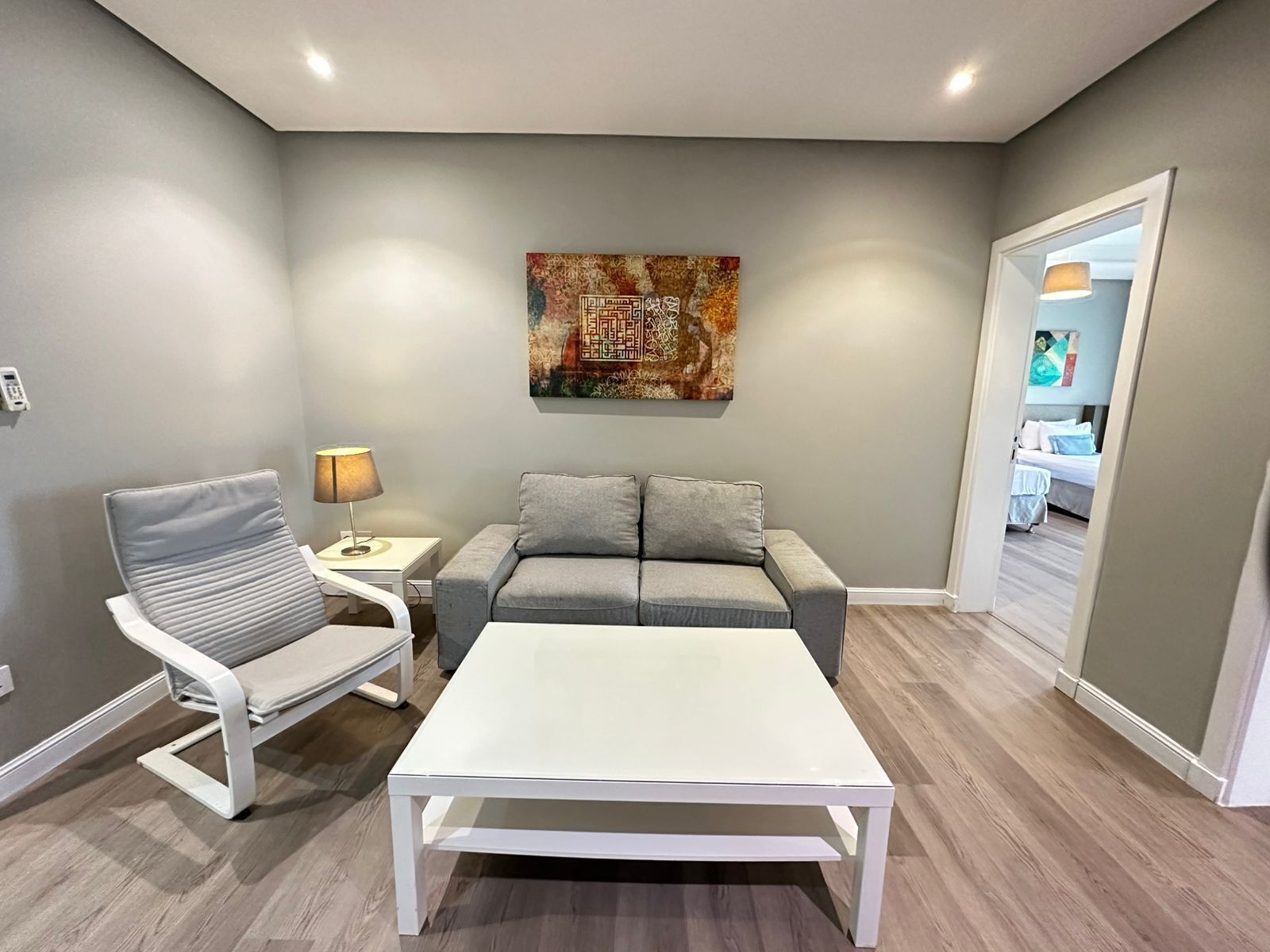 Uma sala de estar moderna com um sofá cinzento, uma mesa de centro branca e uma poltrona cinzenta. Há um quadro na parede e um abajur sobre uma mesinha.