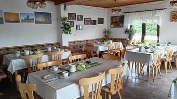 Restaurante com mesas e cadeiras de madeira, toalhas de mesa brancas e plantas em vasos.