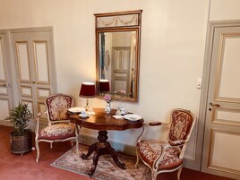 Quarto com mesa redonda de madeira, duas cadeiras decoradas, um espelho e um vaso de planta.