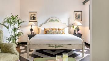 Quarto de hotel com cama, dois mesas de cabeceira, uma cadeira, planta em vaso e mesa com um livro e copos.