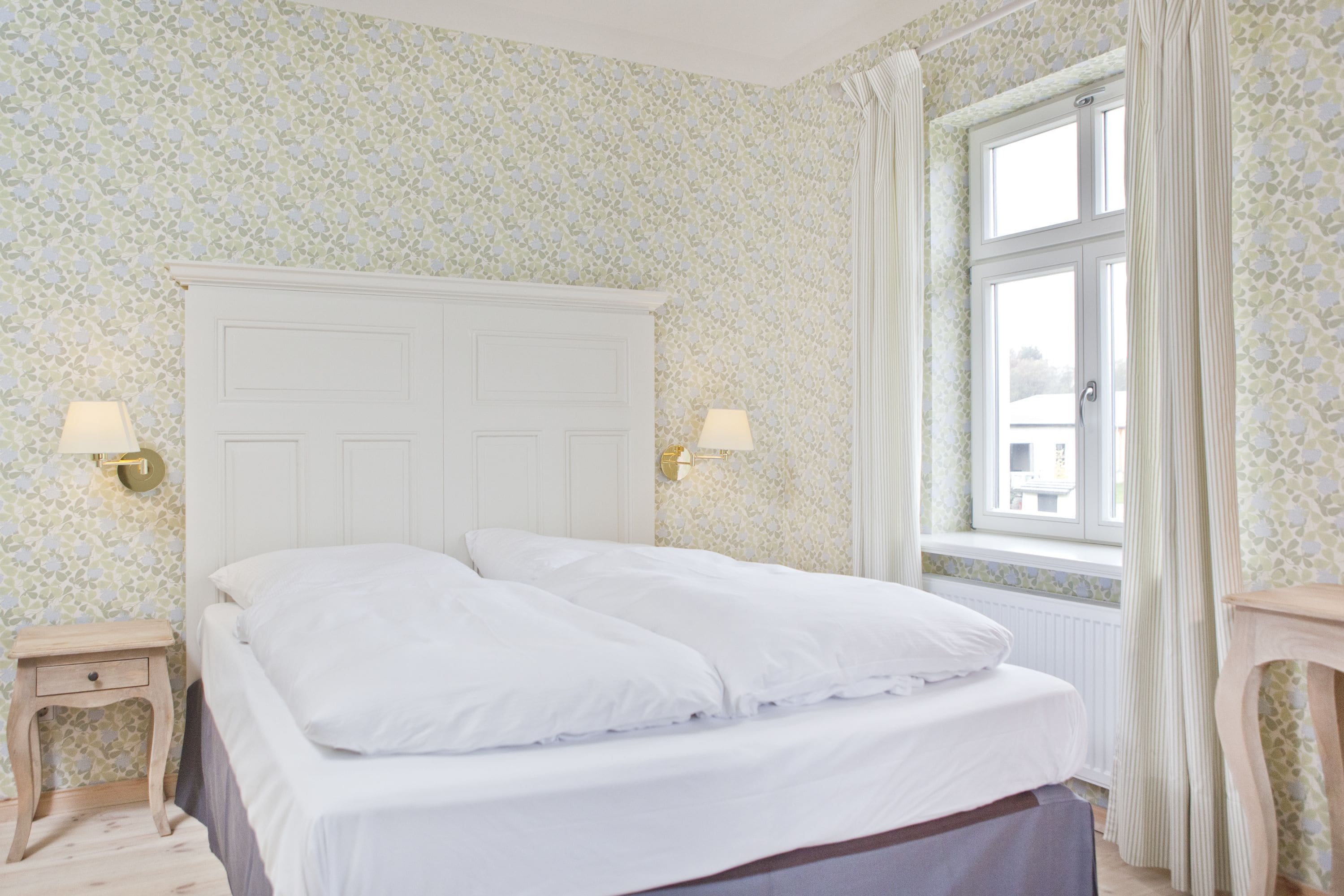 Quarto com cama, duas mesas de cabeceira, janela com cortinas e papel de parede estampado.