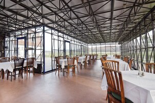 Uma área de refeições interna com mesas e cadeiras, teto alto com vigas de metal e grandes janelas que oferecem vista para o exterior.