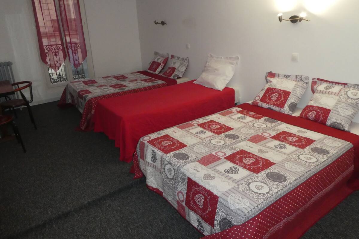 Quarto com duas camas, cada uma com roupa de cama vermelha e colchas estampadas, uma mesinha com cadeiras e uma janela com cortinas.