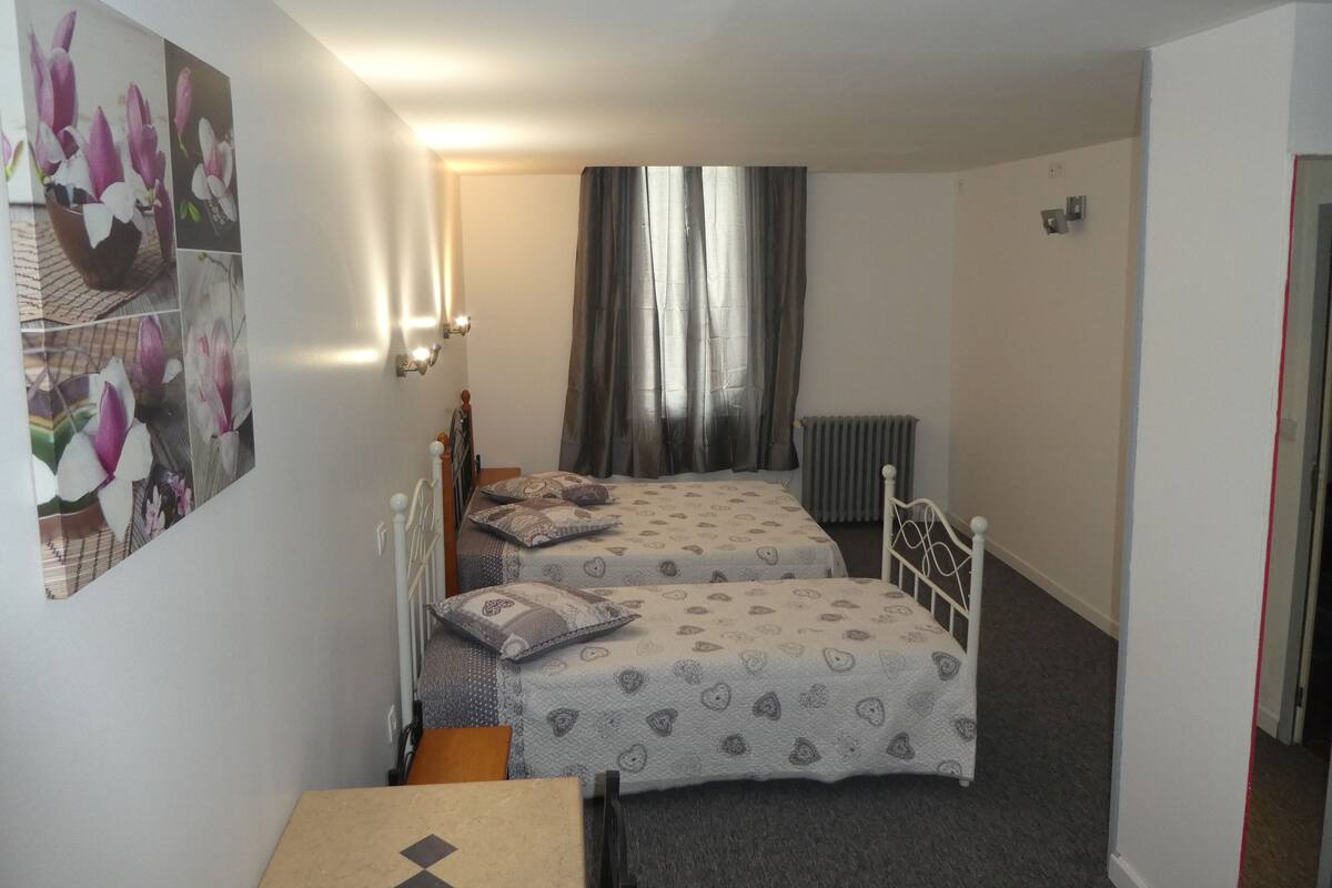 Um quarto pequeno e estreito com duas camas de solteiro, um radiador, uma mesa e um ponto de luz fixado na parede.