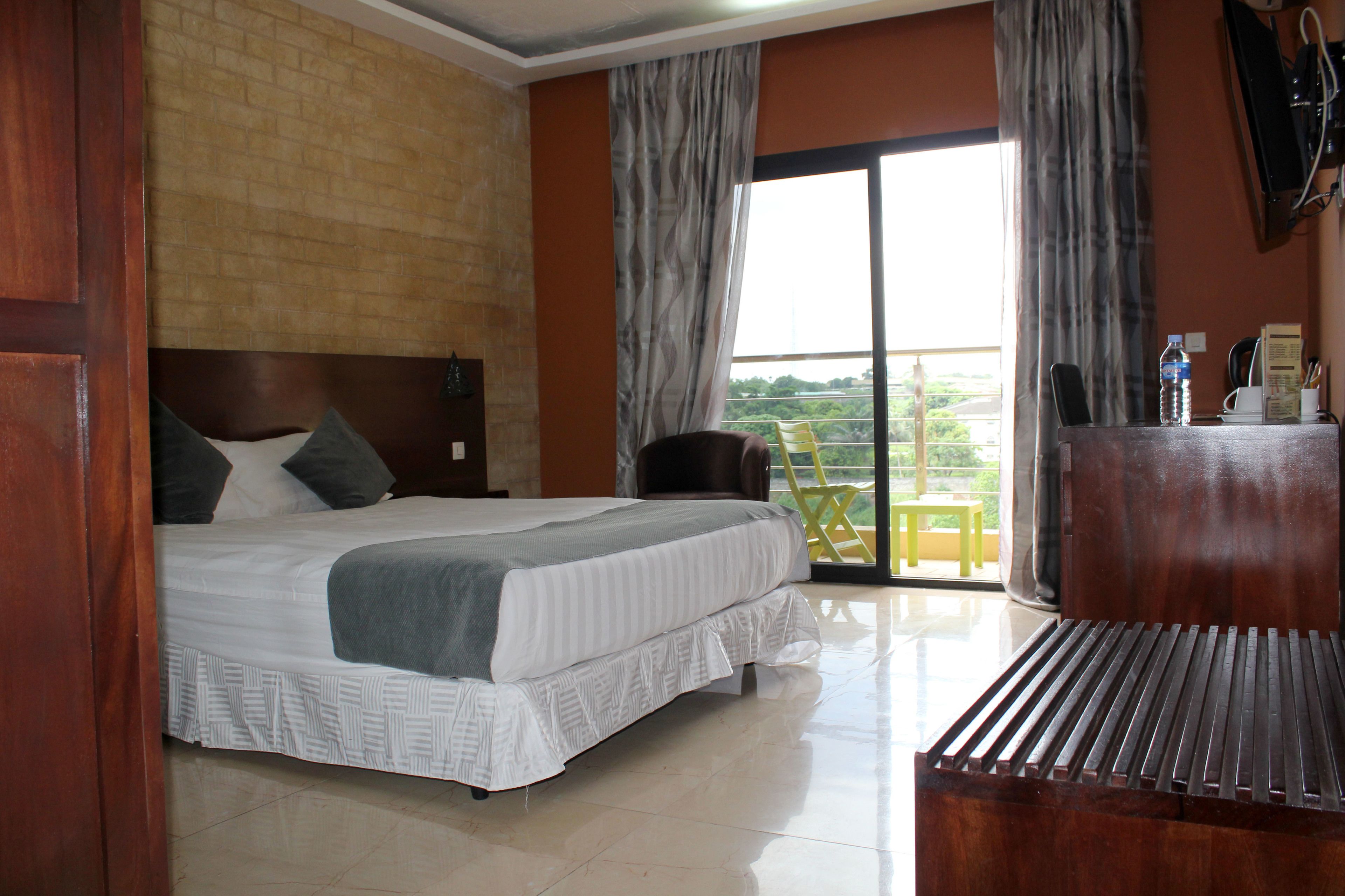 Quarto de hotel com uma cama grande, cabeceira de madeira, escrivaninha com cadeira, janela com cortinas e vista para o verde lá fora.
