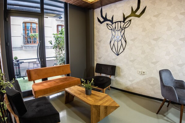 Um interior moderno com um banco de madeira, uma pequena mesa com uma planta, um sofá preto e uma poltrona cinza. É visível uma arte de parede com a cabeça de um veado.
