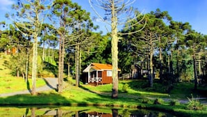 Uma cabana com telhado vermelho e detalhes brancos, rodeada por altas árvores de pinheiro e um pequeno lago.