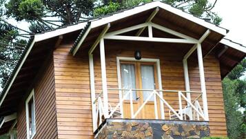 Uma cabana de madeira com alicerce de pedra, varanda com grades brancas e janela com cortinas.
