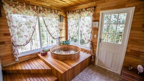 Uma cabana de madeira com banheira de madeira, janelas amplas com cortinas florais e uma porta branca.