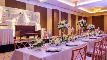 Uma sala de banquetes com longas mesas preparadas para um evento formal, com toalhas brancas, arranjos florais e elegantes luminárias.