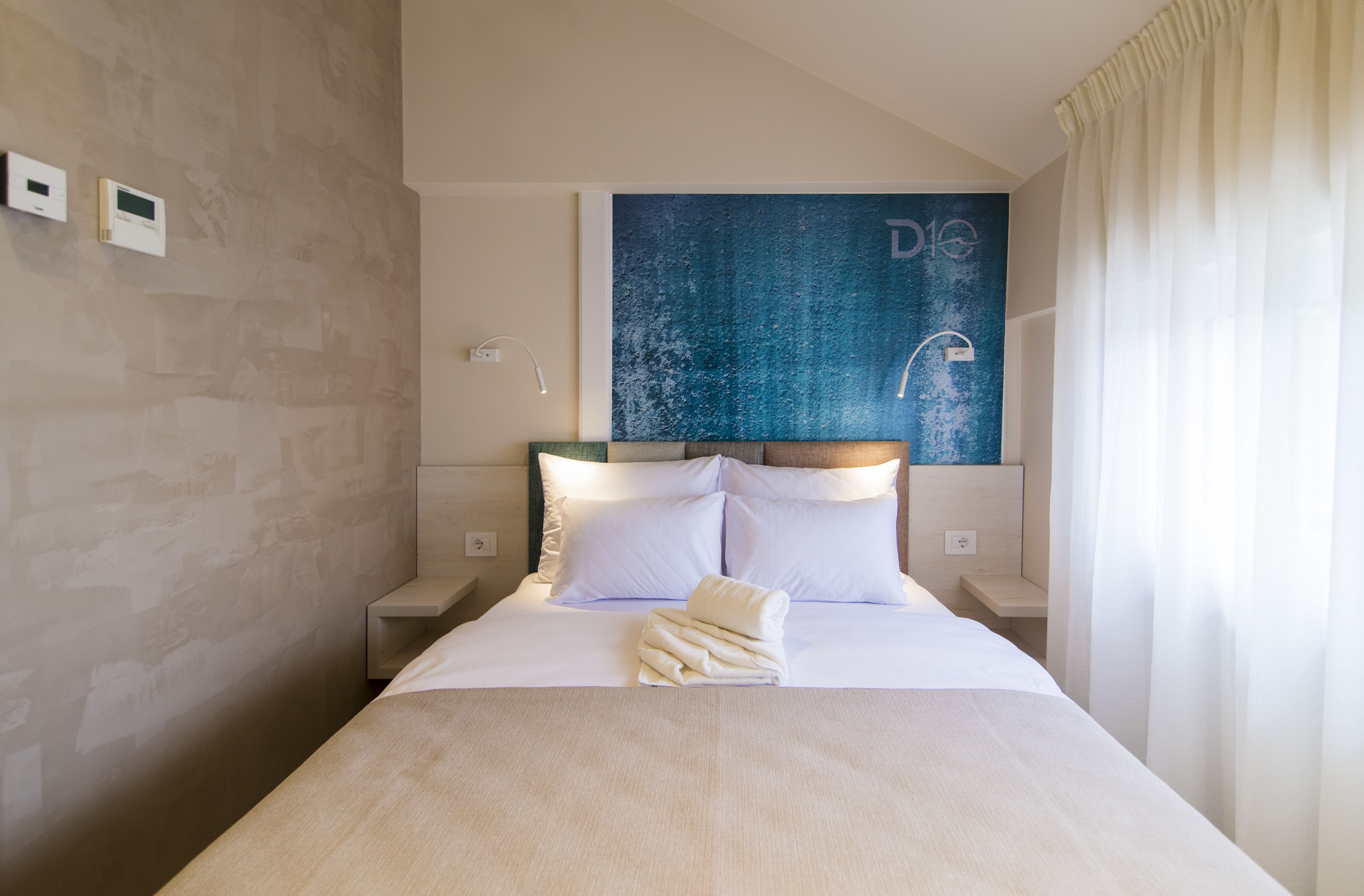 Quarto de hotel com uma cama grande, duas mesas de cabeceira, uma obra de arte na parede e uma janela com cortinas.