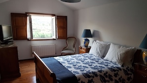 Quarto com cama, poltrona, mesa de cabeceira, abajur, cômoda, janela e radiador.