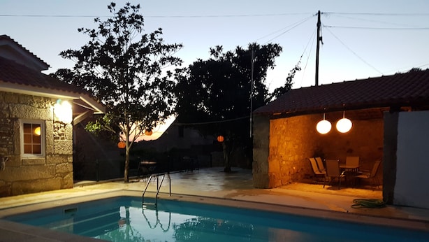 Piscina com escada, cercada por paredes de pedra e uma árvore. Há cadeiras e mesa ao ar livre sob um candeeiro.