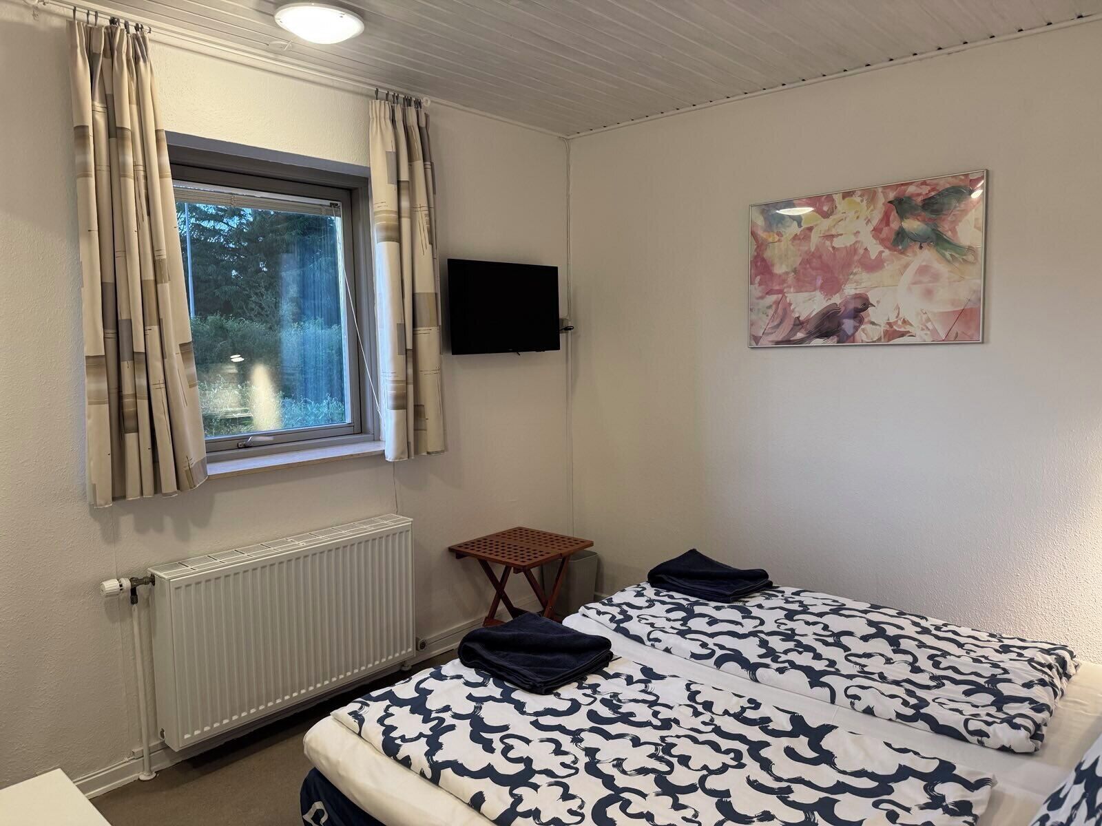 Quarto com duas camas, janela com cortinas, radiador, mesinha e um quadro na parede.