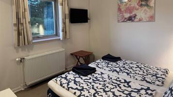 Quarto com duas camas, janela com cortinas, radiador, mesinha e um quadro na parede.