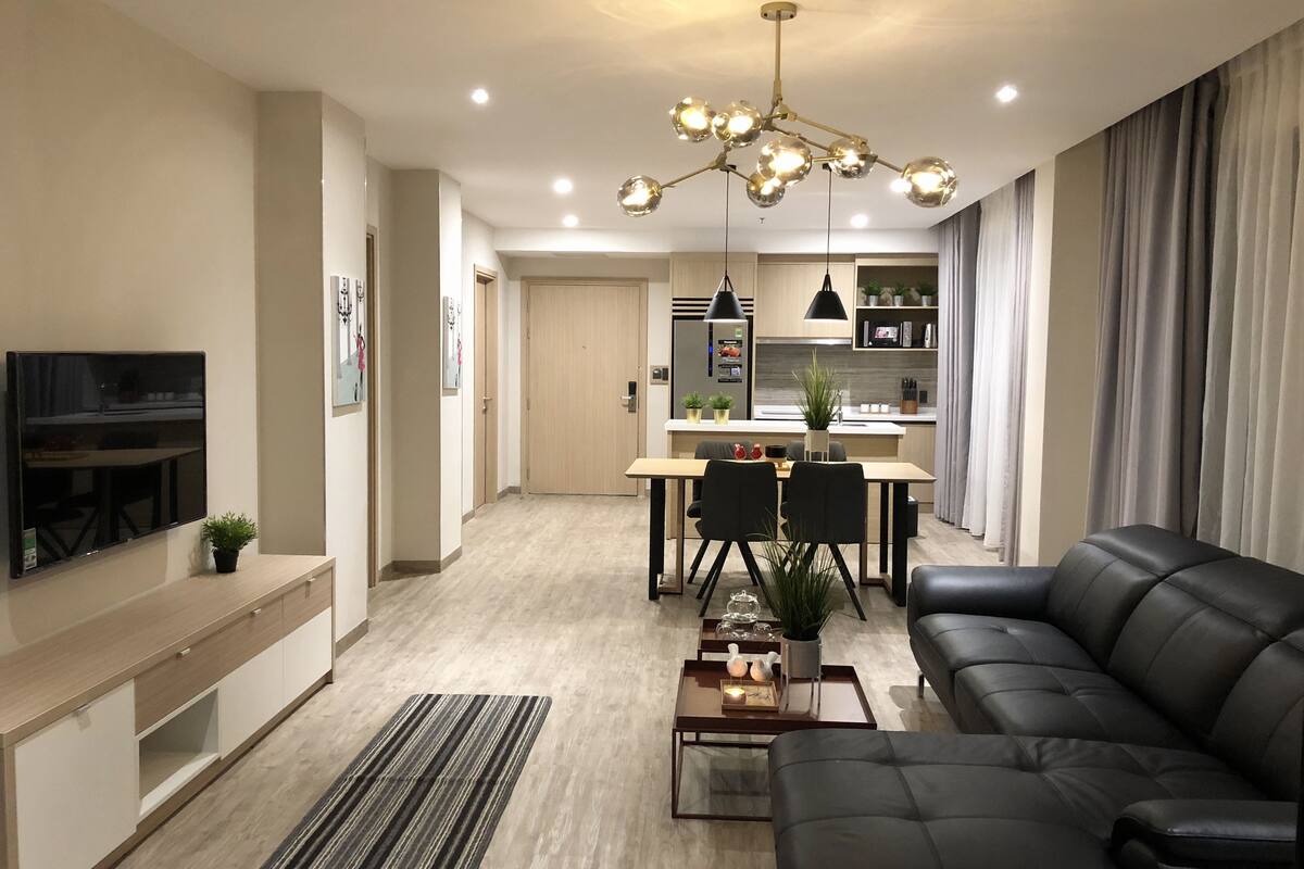 Uma sala de estar moderna com um sofá de couro preto, uma mesa de centro de madeira e uma TV fixada na parede. Há uma área de jantar com mesa e cadeiras, e uma cozinha ao fundo.