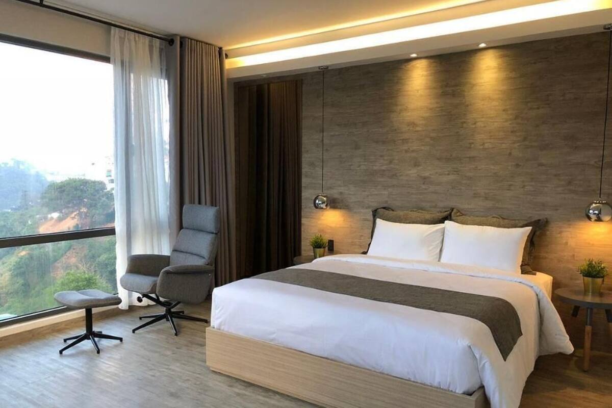Quarto moderno com cama grande, escrivaninha e cadeira, vista para o exterior e parede de madeira.