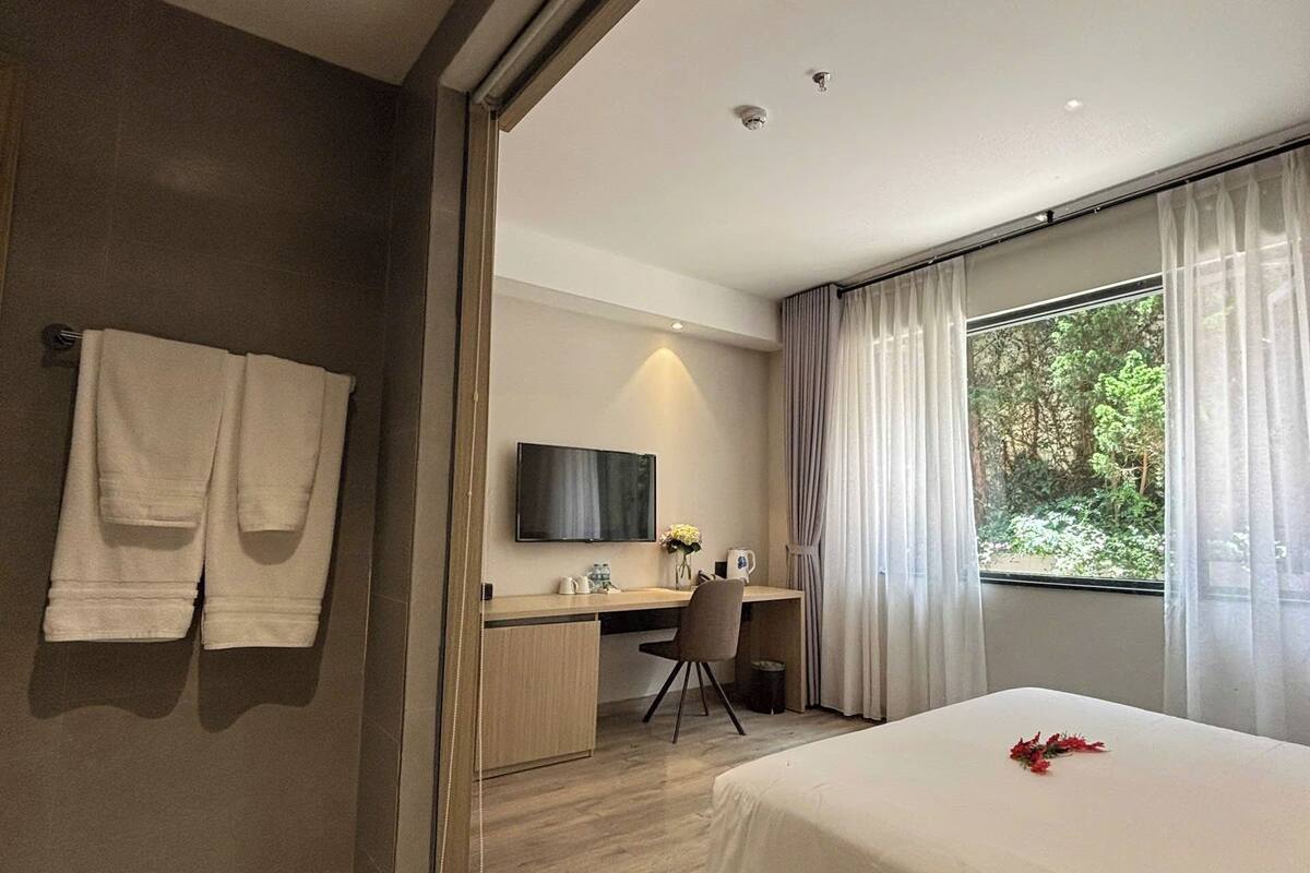 Quarto de hotel moderno com cama, escrivaninha, cadeira, televisão e banheiro com toalhas.
