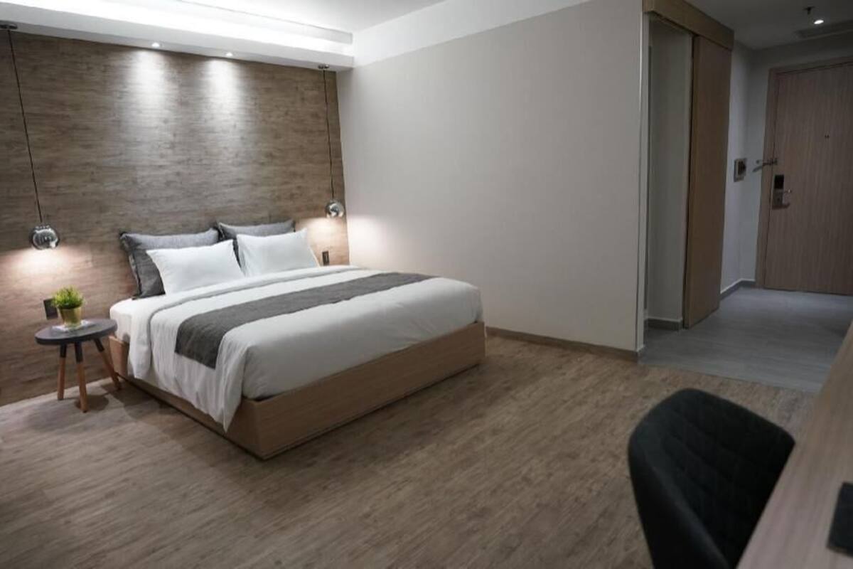 Quarto de hotel moderno com uma cama grande, mesas de cabeceira, uma pequena planta e uma porta que dá para outro quarto.
