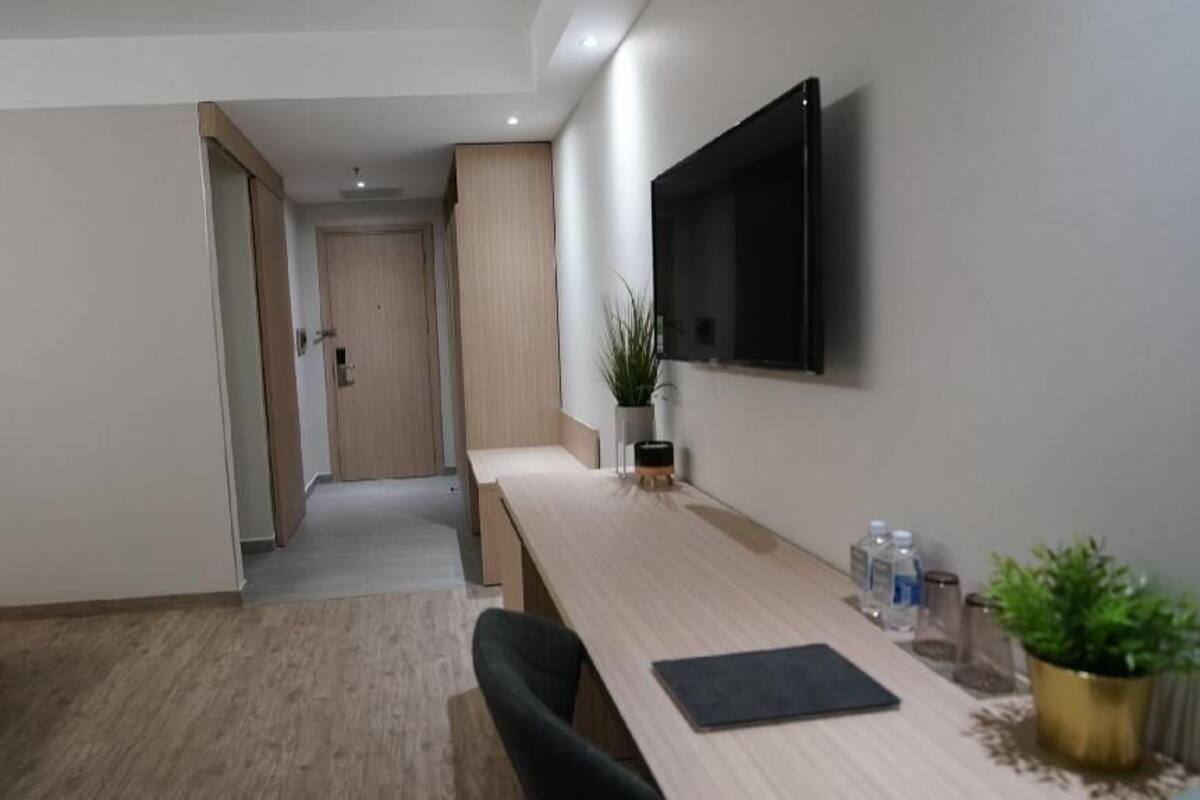 Quarto de hotel com cama, escrivaninha com cadeira, televisão na parede e planta em vaso.