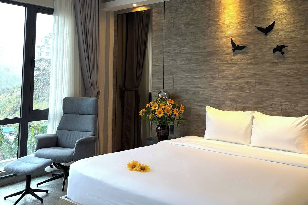 Quarto de hotel moderno com uma cama grande, uma poltrona, uma escrivaninha e vista para o exterior.