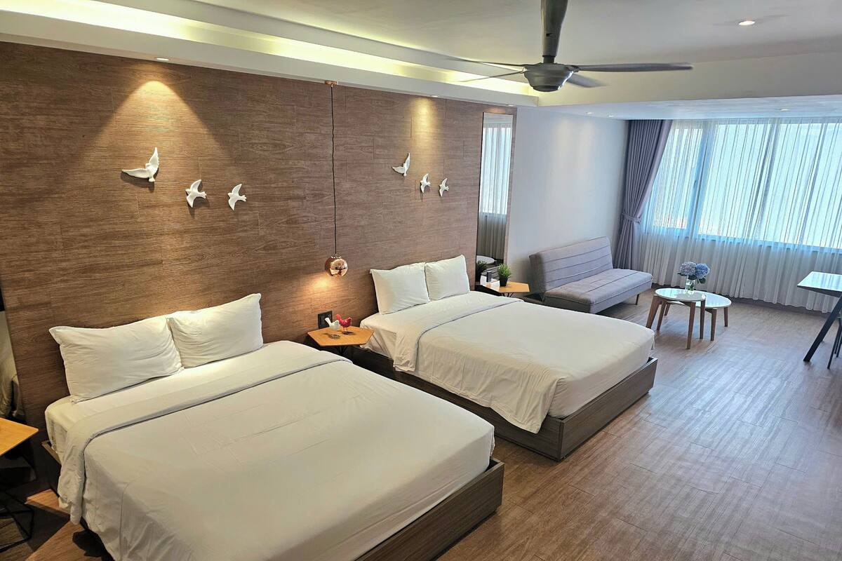 Quarto de hotel com duas camas, um ventilador de teto, uma mesinha com uma planta e um sofá.