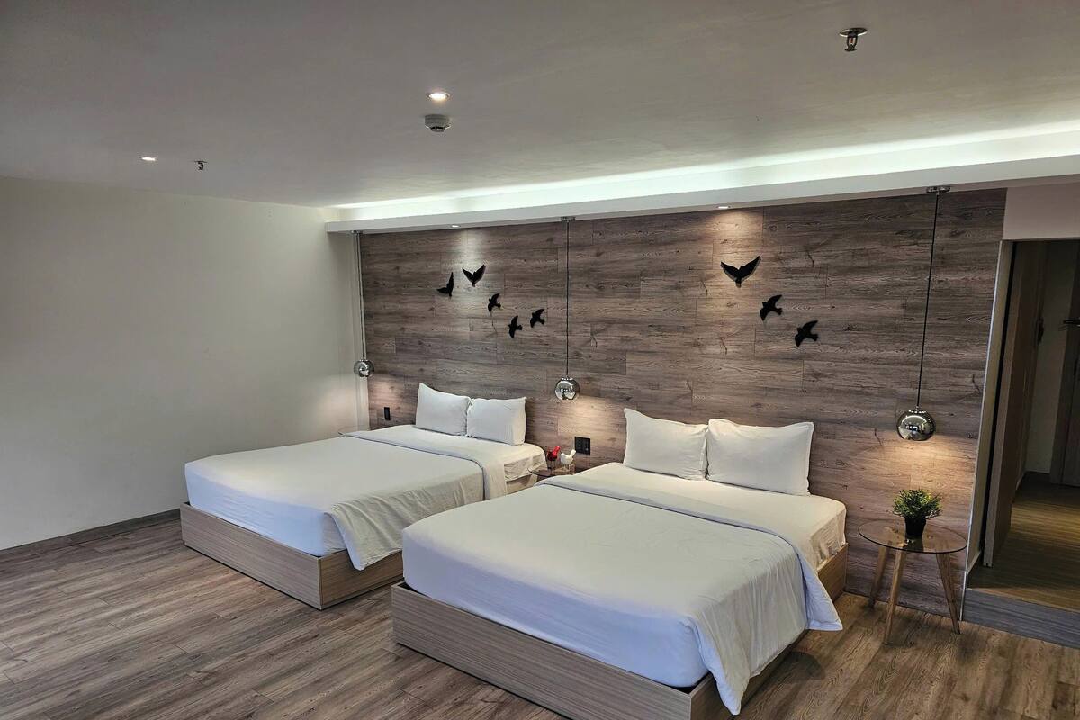 Quarto de hotel moderno com duas camas, piso de madeira, painel de parede em madeira e luminária suspensa no teto.