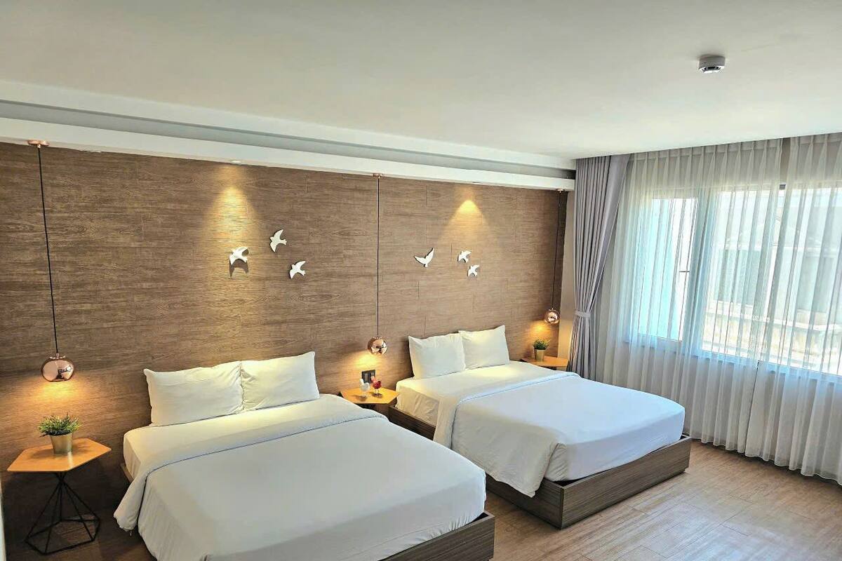 Quarto de hotel moderno com duas camas, cabeceira de madeira, luminárias fixadas na parede e uma janela grande com cortinas translúcidas.