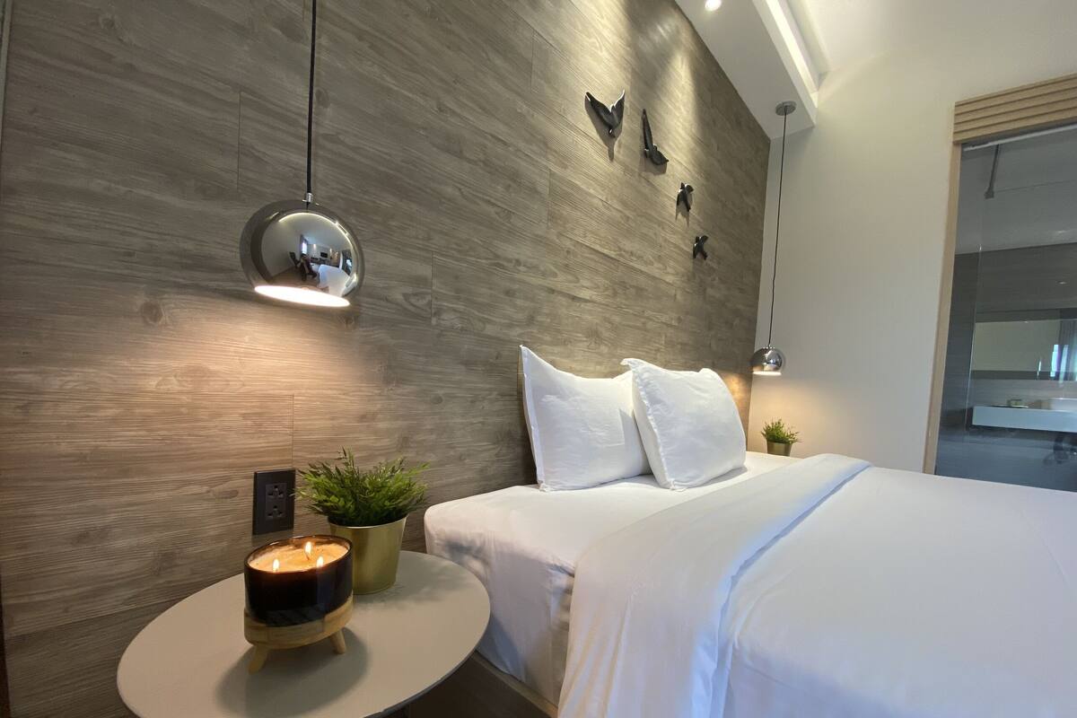 Quarto moderno com parede de madeira, mesa redonda com vela e cama com roupa de cama branca.