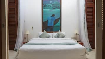 Uma cama grande com roupa de cama branca e almofadas listradas, um quadro de uma pessoa num barco, e persianas de madeira nas janelas.