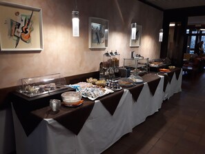 Disposição de buffet com diversos alimentos, incluindo doces e sobremesas, dispostos sobre uma mesa coberta com toalha branca.