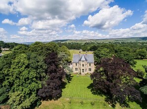 Um edifício grande e histórico com fachada simétrica, rodeado por vegetação exuberante e um gramado bem cuidado.