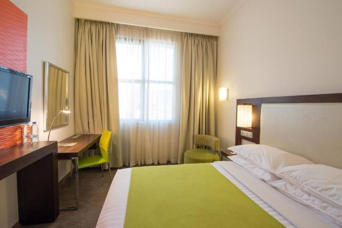 Quarto de hotel com uma cama, uma escrivaninha com cadeira, televisão e uma janela com cortinas.