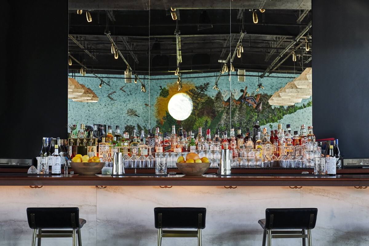 Um bar com balcão extenso, diversas garrafas, copos e um mural ao fundo.