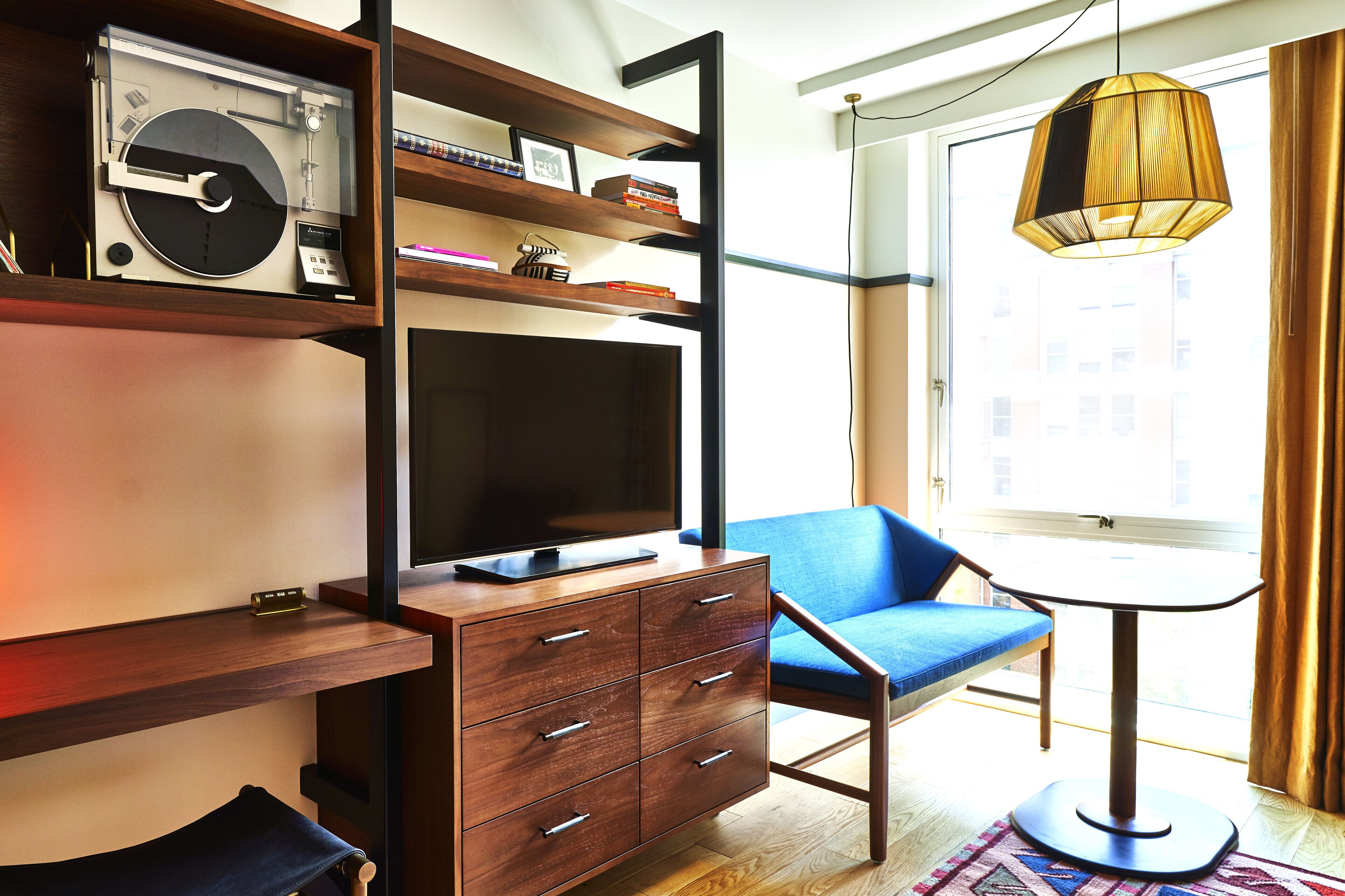 Quarto moderno com televisão sobre um armário de madeira, uma cadeira azul e uma pequena mesa com abajur.