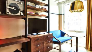 Quarto moderno com televisão sobre um armário de madeira, uma cadeira azul e uma pequena mesa com abajur.