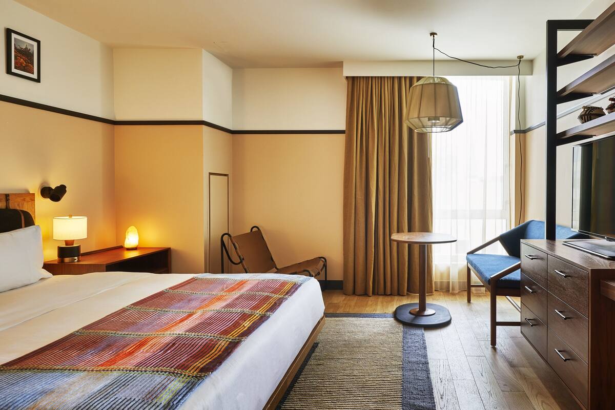 Quarto de hotel com cama, escrivaninha, cadeira, luminária, televisão e janela com cortinas.