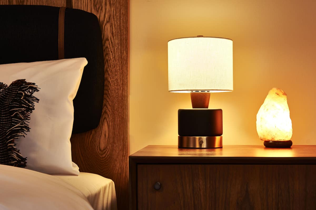 Uma cama com almofada branca e almofada decorativa com franjas. Um criado-mudo de madeira com abajur e uma luminária de sal fica ao lado da cama.