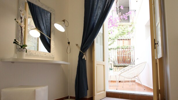 Casa de banho com banheira branca, espelho, candeeiro de chão e vista para uma varanda exterior com uma cadeira.